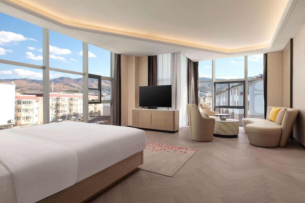 Quarto de hotel moderno com uma cama grande, televisão, área de estar com mesa e grandes janelas com vista para a cidade.