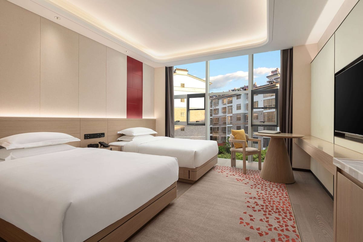 Quarto de hotel moderno com duas camas, televisão, mesa pequena e varanda com vista para a cidade.