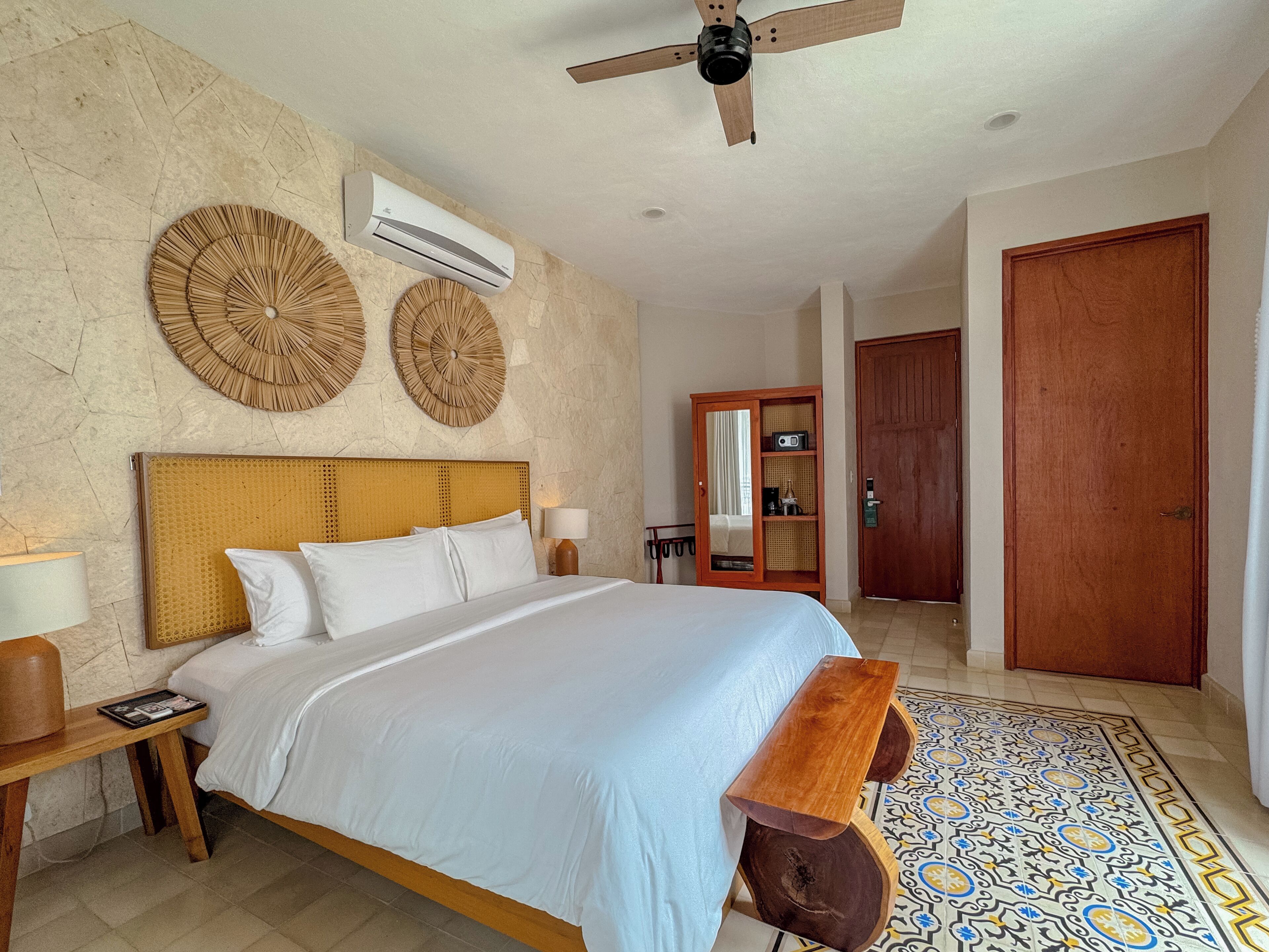 Quarto com cama, mesa de cabeceira, abajur, ar condicionado, ventoinha de teto e um banco de madeira.