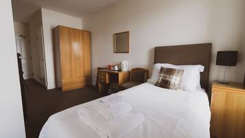 Quarto de hotel com uma cama grande, um guarda-roupa de madeira, uma escrivaninha com cadeira, mesa de cabeceira com abajur e espelho na parede.