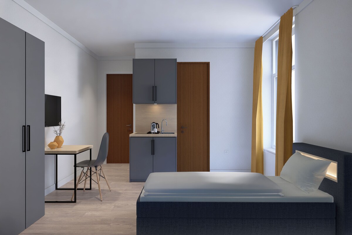 Quarto de hotel moderno com cama, escrivaninha com cadeira, cozinha compacta e televisão.