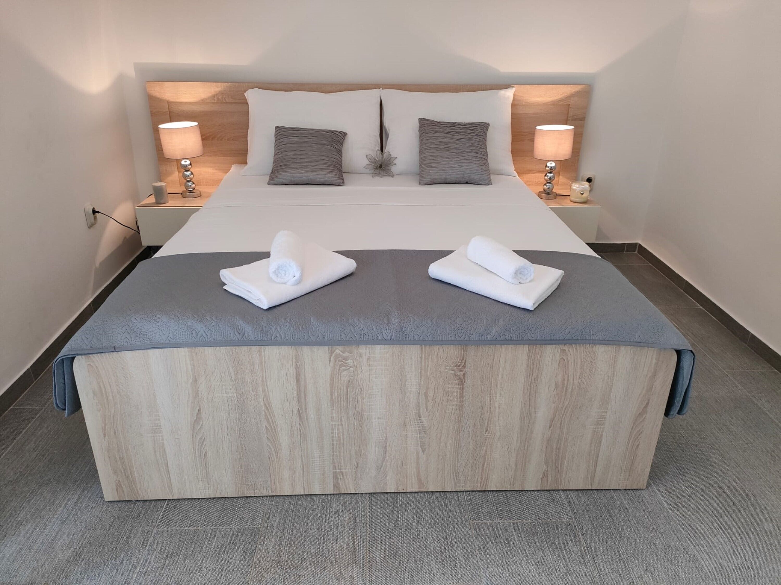 Cama bem arrumada com duas toalhas brancas, cabeceira de madeira e duas luminárias de apoio.