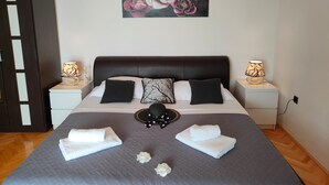 Cama bem arrumada com um edredom cinza, duas toalhas brancas e uma almofada decorativa em preto e branco. Há uma pintura de flores na parede acima da cama.