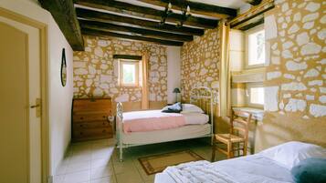 Quarto com parede de pedra, cama de madeira, cômoda de madeira, uma cadeira e uma janela.