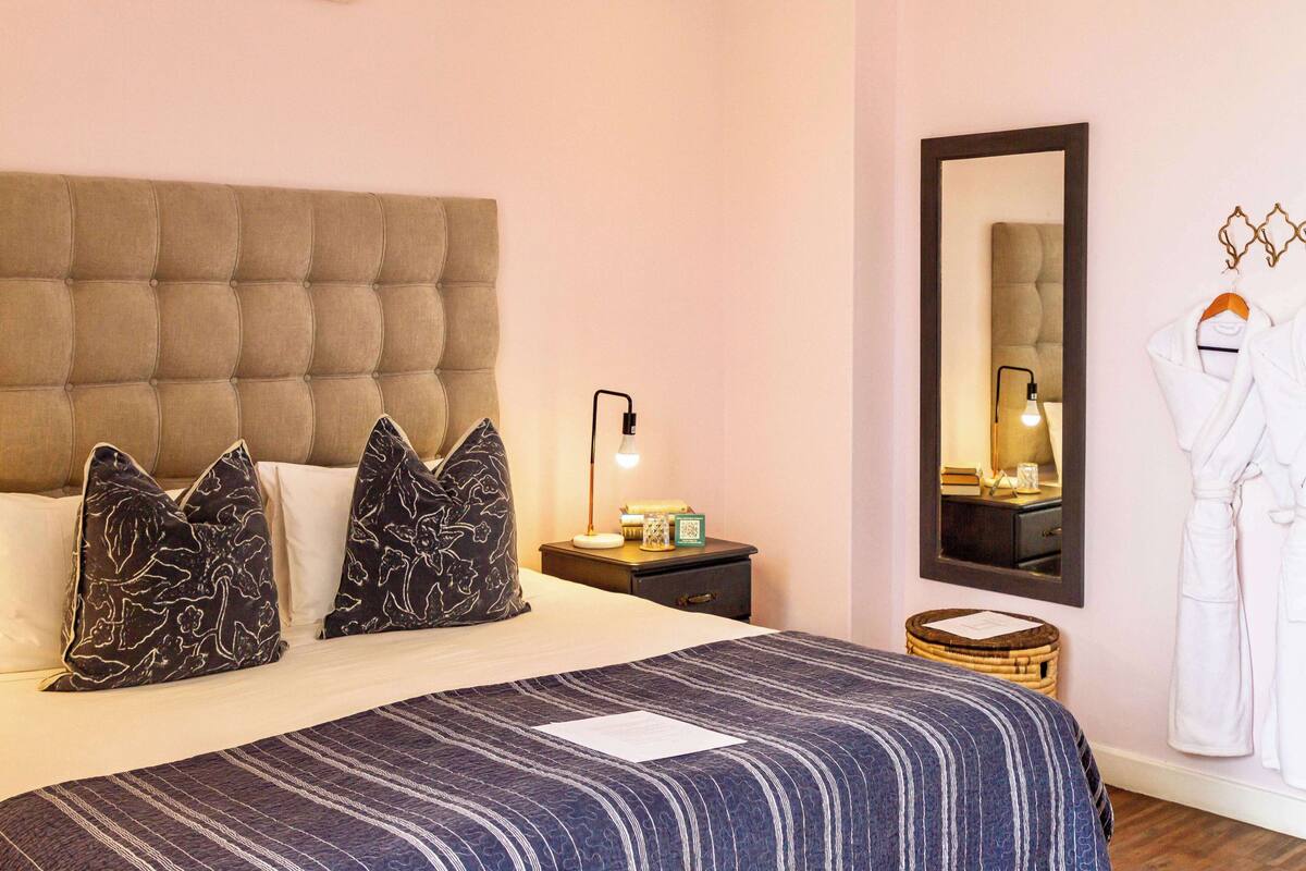 Cama bem arrumada com colcha listrada, duas almofadas decorativas, abajur, mesa de cabeceira pequena com luminária, espelho e roupão pendurado na parede.