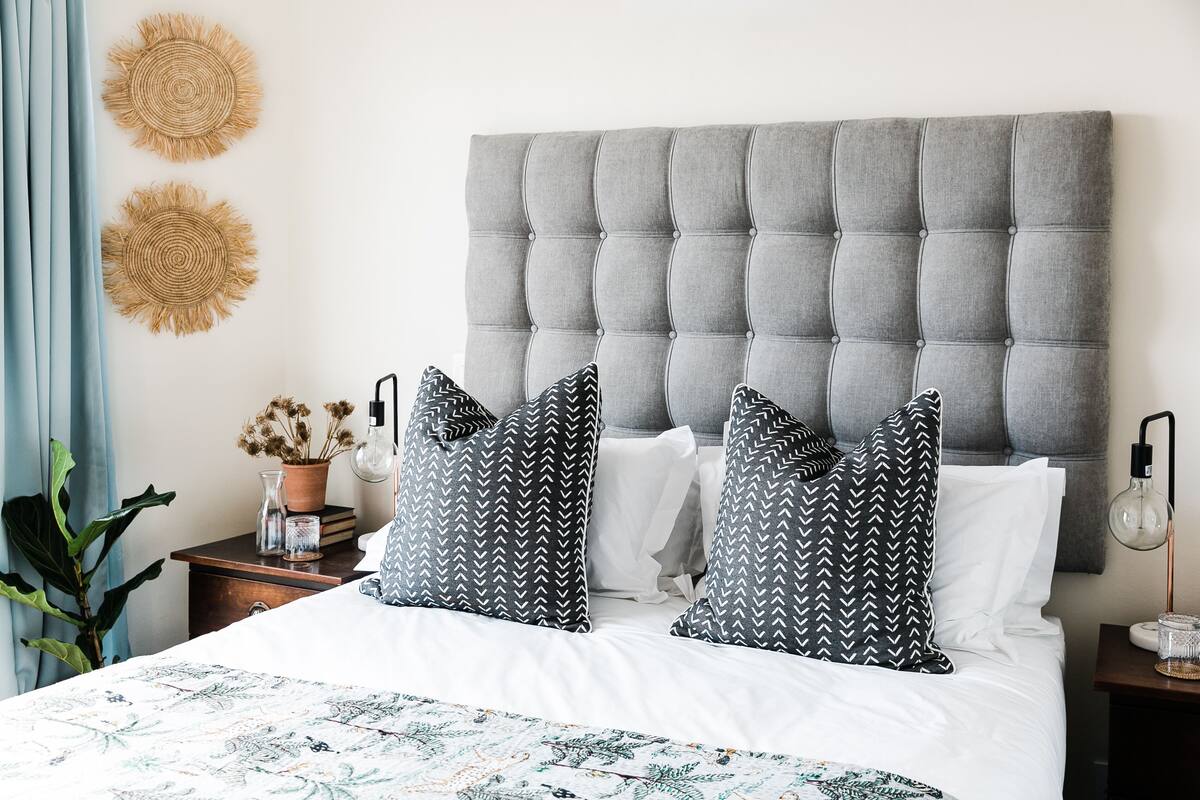 Cama bem arrumada com cabeceira estofada cinza, roupa de cama branca e almofadas estampadas.