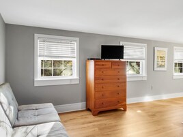 Um quarto com parede cinza, piso de madeira, sofá cinza, cômoda de madeira e janelas com moldura branca e persianas.