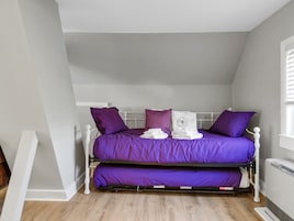 Uma cama beliche de dois níveis com roupa de cama roxa, cabeceira branca e um radiador branco na parede.