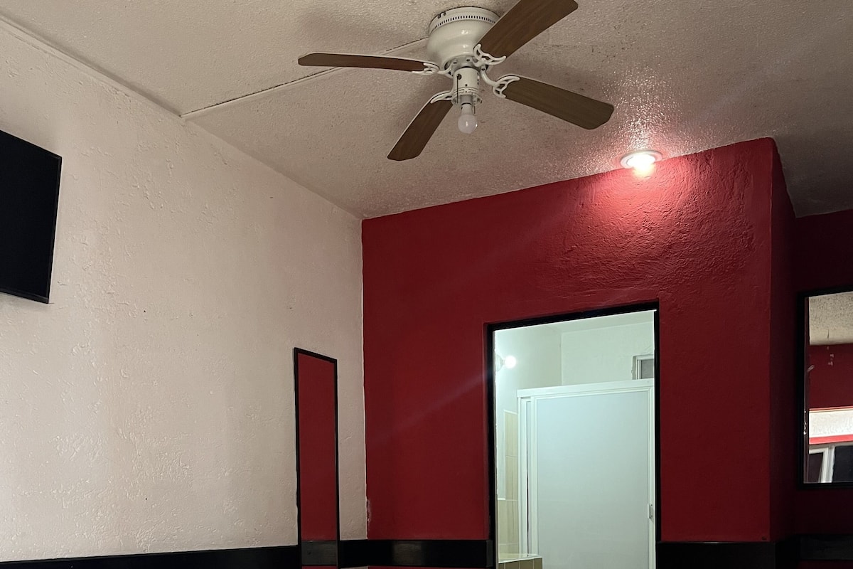 Quarto com cama, ventilador de teto, parede de destaque vermelha, porta e espelho.