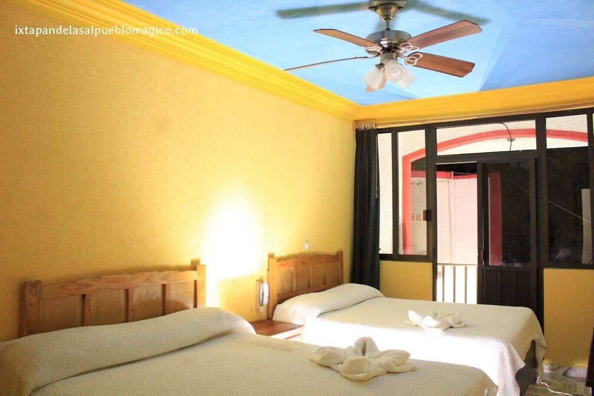Quarto de hotel com duas camas, ventilador de teto e janela com vista para o exterior.