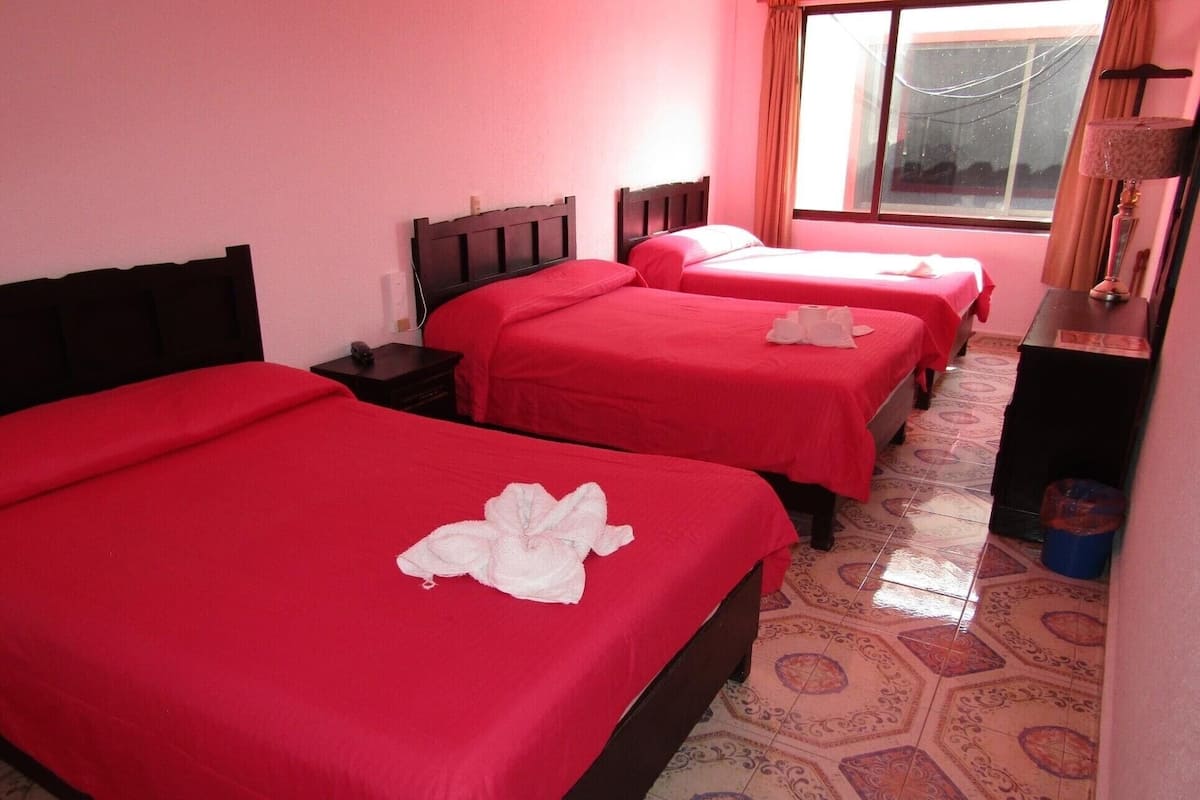 Quarto de hotel com três camas, cada uma com colcha vermelha e toalhas brancas. O quarto possui ladrilhos no chão com padrão e uma janela com cortinas.