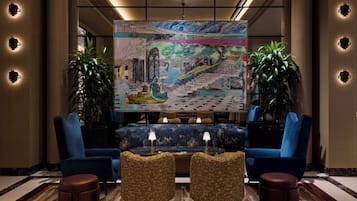 Um hall de entrada com uma grande pintura abstrata, poltronas estofadas em azul, um sofá e duas mesas com detalhes dourados.