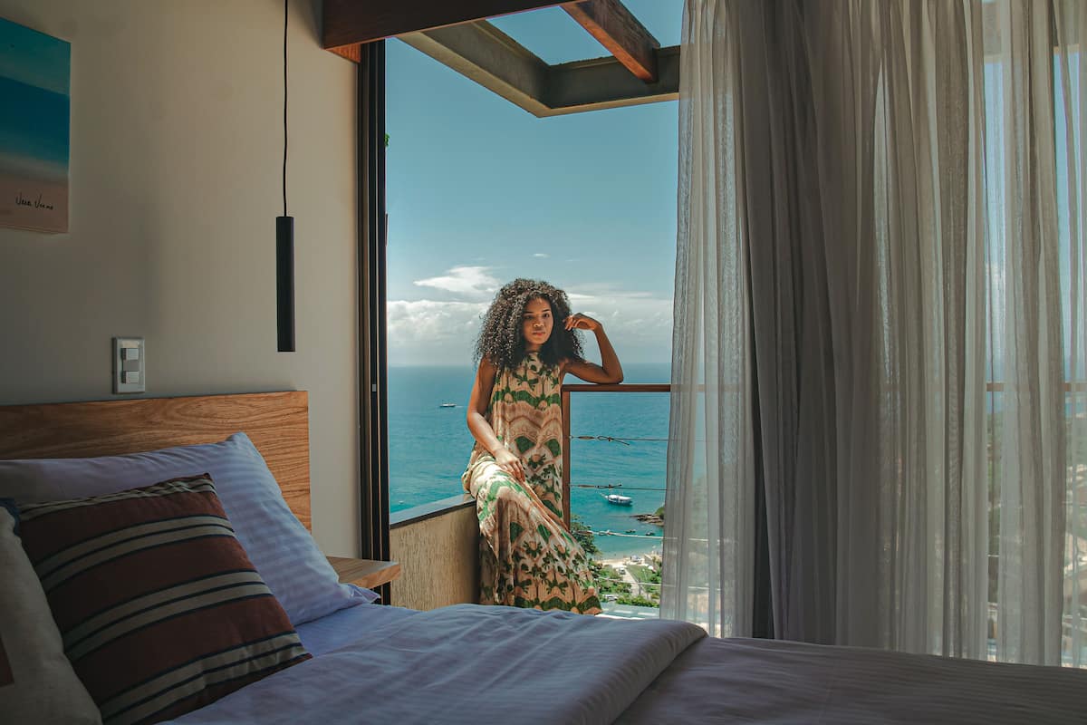 Quarto com cama grande, vista para o mar e varanda com uma pessoa.
