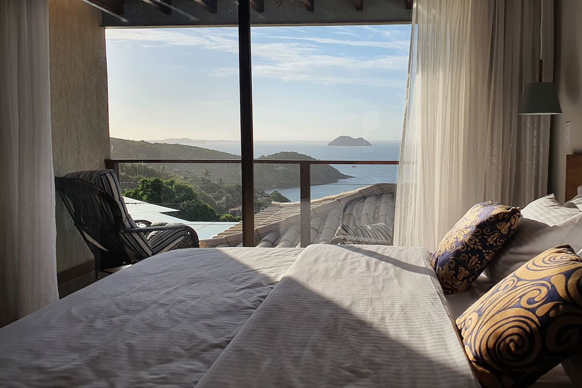 Quarto com cama grande, uma cadeira e vista para o oceano.