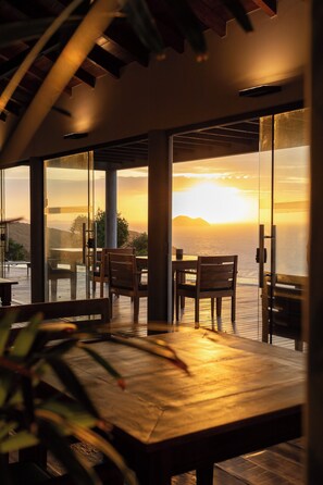 Uma área de refeições com vista para o pôr do sol sobre o oceano, com uma mesa e cadeiras de madeira.