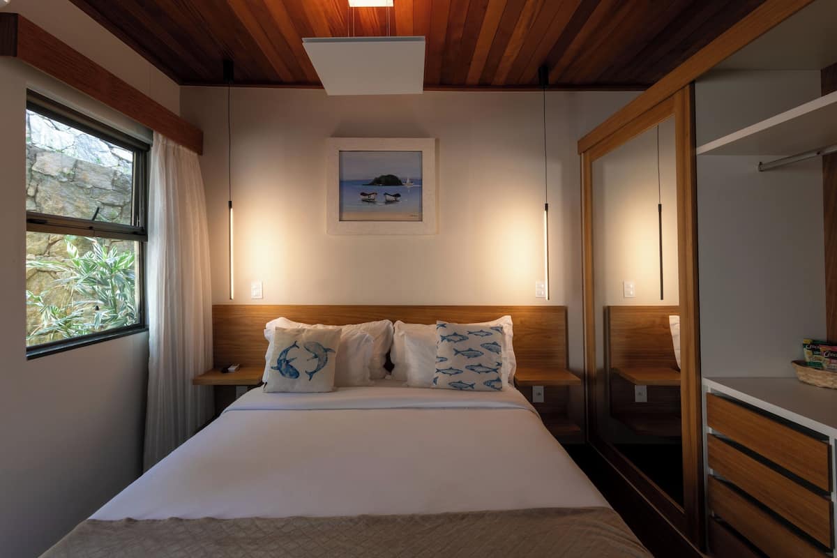Cama bem arrumada com edredom branco e almofadas com padrão de peixes azuis, cabeceira de madeira, quadro emoldurado acima da cama e janela com cortinas à esquerda.