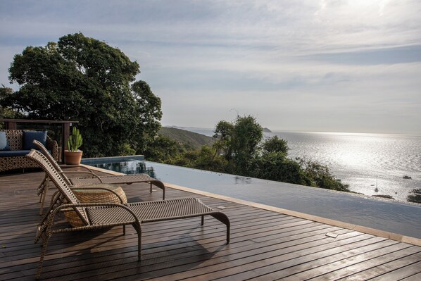 Um deck de madeira com cadeiras de lounge de vime, com vista para um corpo de água e uma paisagem distante.