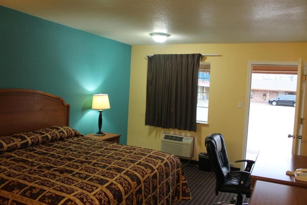 Quarto de hotel com cama, escrivaninha, cadeira, luminária, janela com cortinas e ar condicionado.