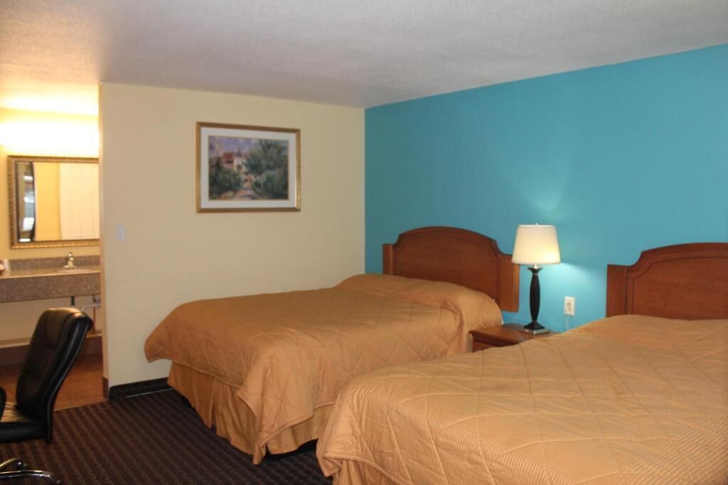 Quarto de hotel com duas camas, uma escrivaninha, uma cadeira, um abajur e um quadro na parede.