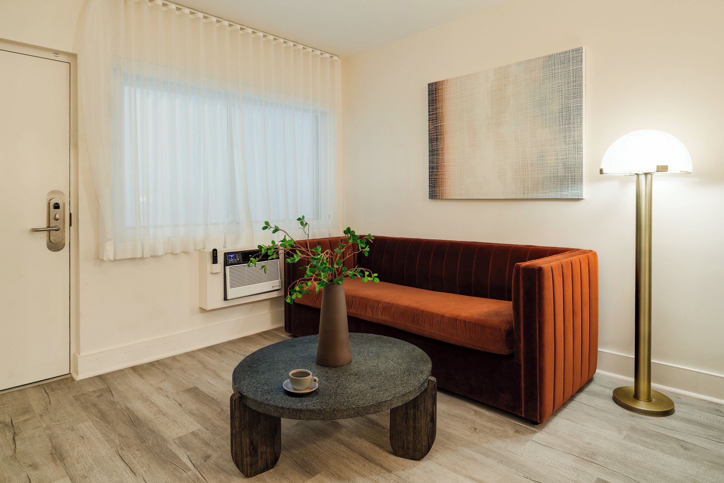 Quarto com sofá marrom, mesa redonda com vaso e chávena, candeeiro de chão, quadro na parede e janela com cortinas.