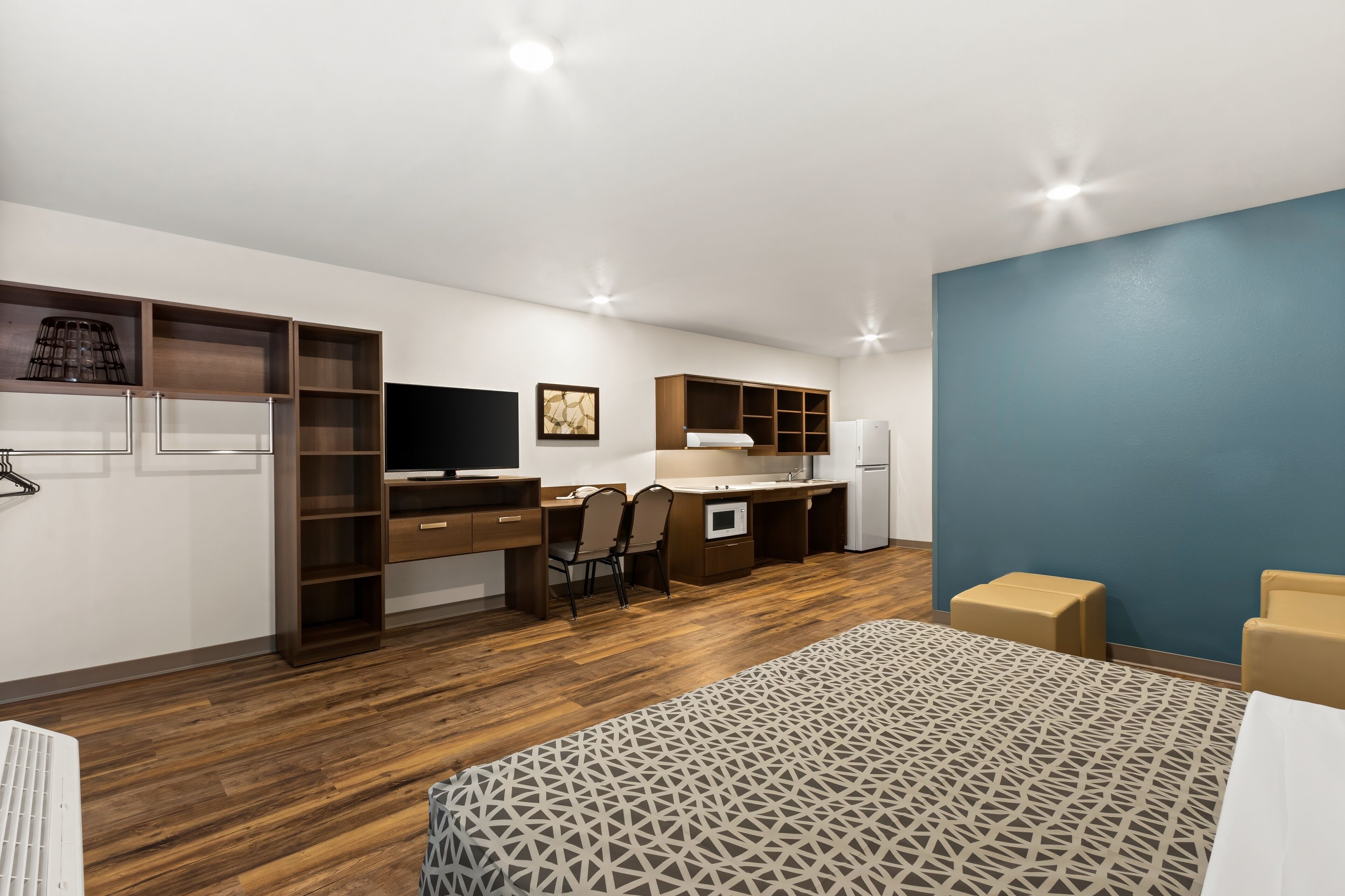 Quarto de hotel moderno com piso de madeira, cama com colcha de padrão geométrico, escrivaninha com computador, cadeira, pequena cozinha compacta e prateleiras embutidas.