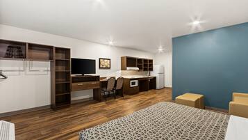 Quarto de hotel moderno com piso de madeira, cama com colcha de padrão geométrico, escrivaninha com computador, cadeira, pequena cozinha compacta e prateleiras embutidas.