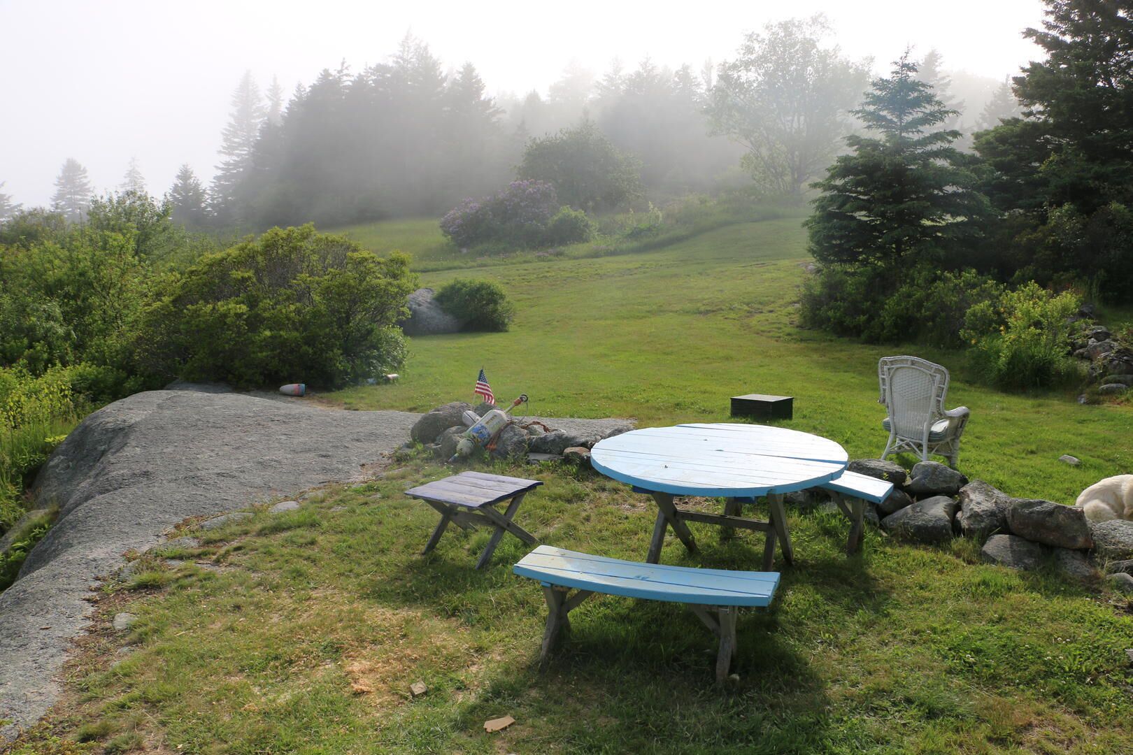 Uma área de estar rústica ao ar livre, com uma mesa redonda e duas cadeiras, cercada por pedras e vegetação.