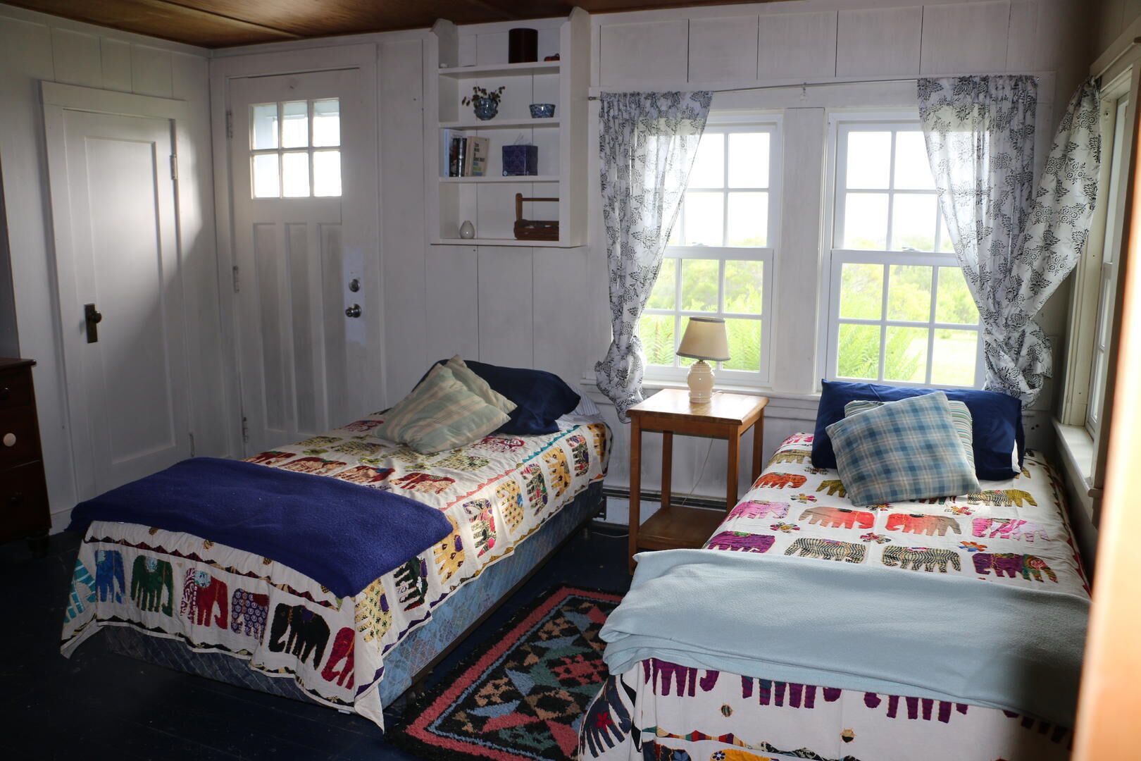Um quarto acolhedor com duas camas, uma mesinha de cabeceira com abajur e uma janela com cortinas.