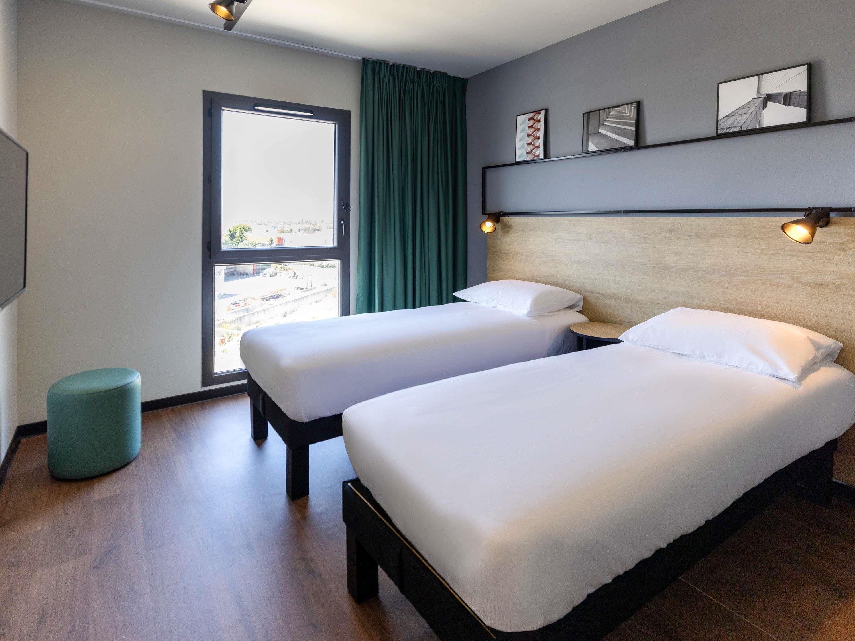 Quarto de hotel com duas camas, uma janela com cortinas, um banquinho e uma obra de arte na parede.