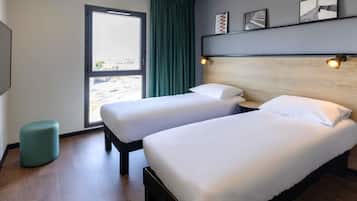 Quarto de hotel com duas camas, uma janela com cortinas, um banquinho e uma obra de arte na parede.