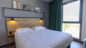 Cama bem arrumada com lençóis brancos, cabeceira com duas luzes fixadas na parede e janela com cortinas verdes.