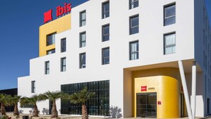 Um hotel moderno com um logotipo 'ibis' vermelho em destaque na fachada, várias janelas e uma parede de detalhes em amarelo.
