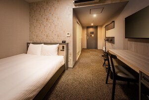 Quarto de hotel com uma cama, uma escrivaninha e uma cadeira. Há uma porta que dá para outro quarto e uma televisão fixada na parede.