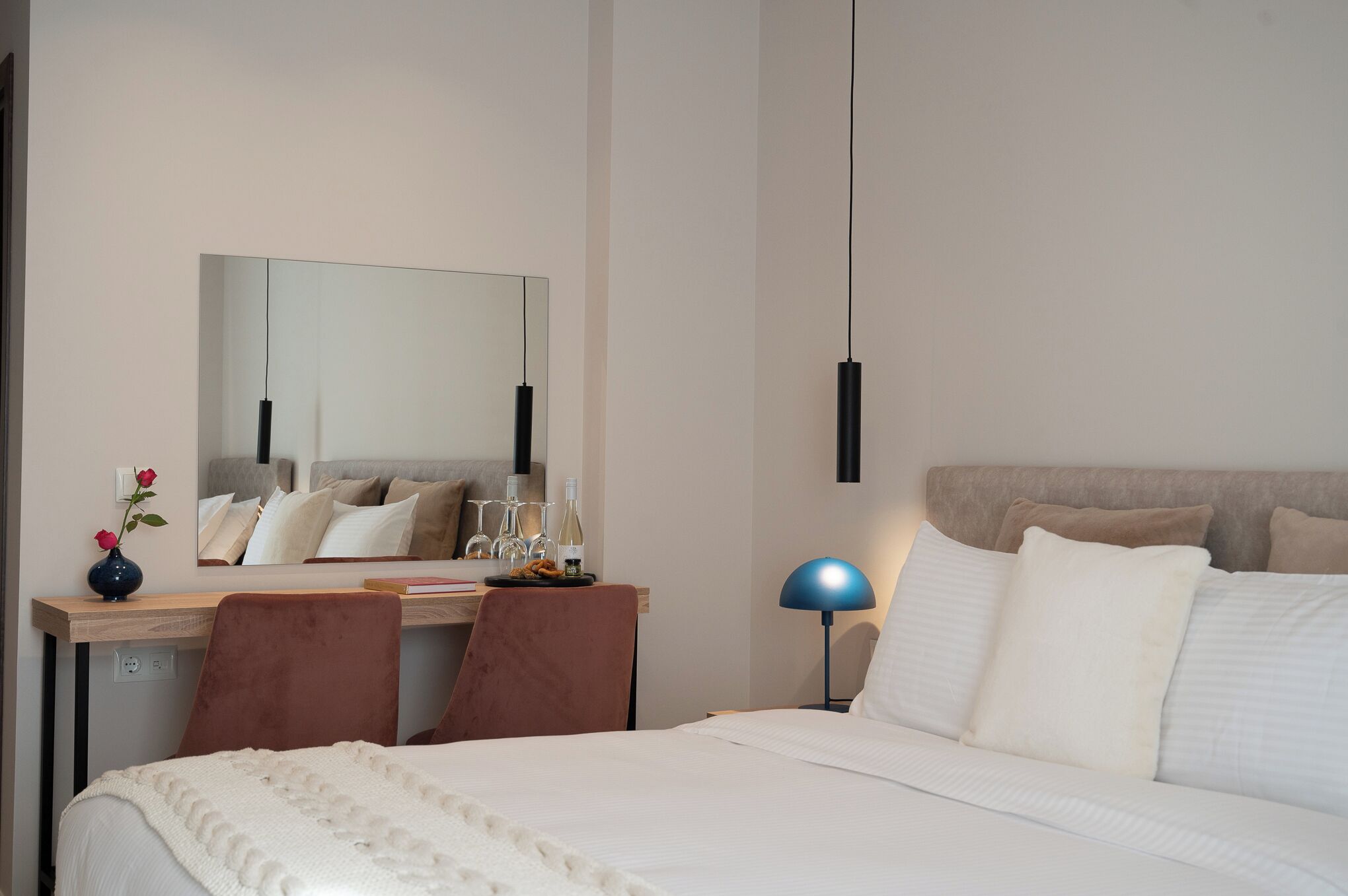 Quarto de hotel moderno com uma cama grande, uma escrivaninha com cadeira, um espelho e uma mesa de cabeceira com abajur.