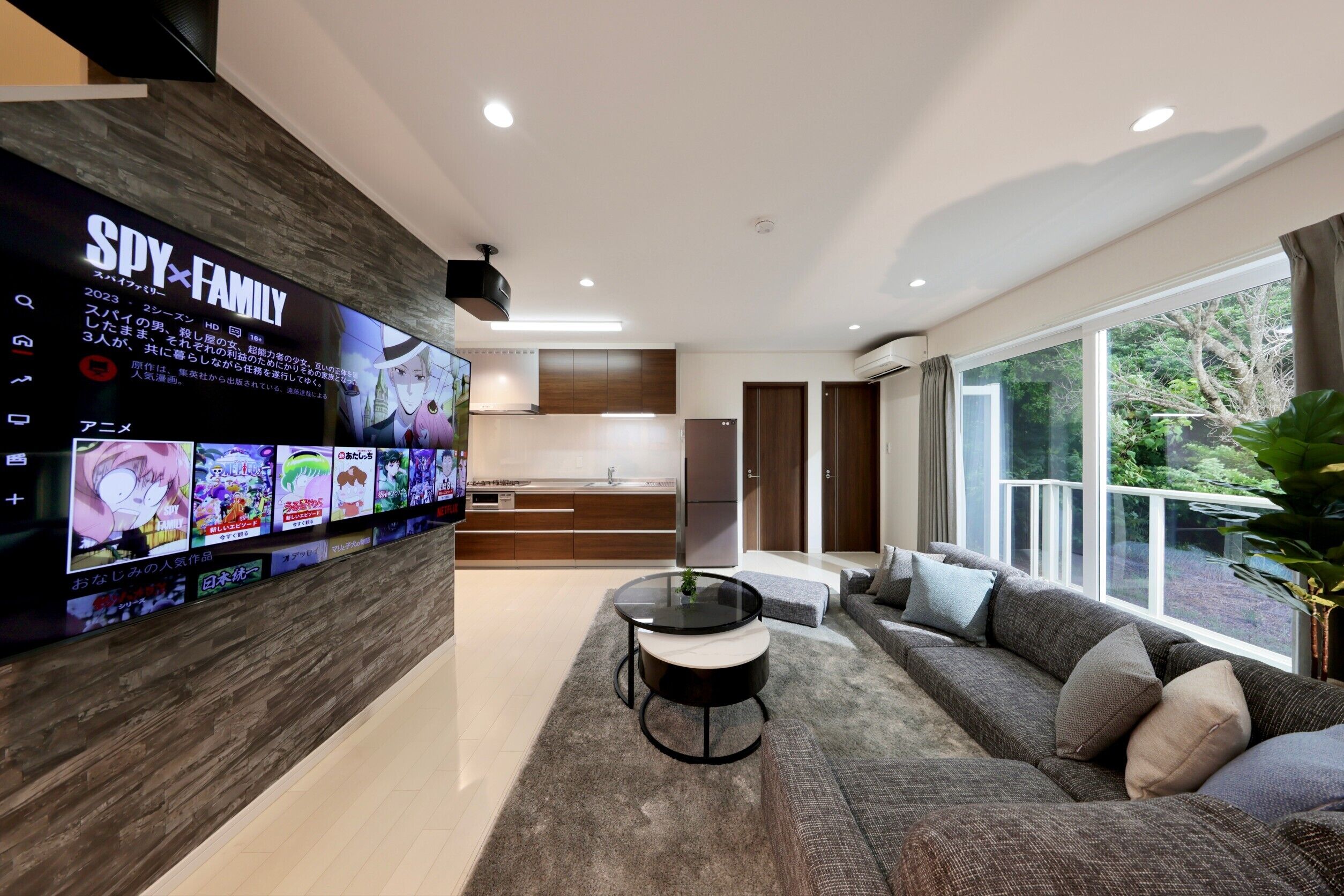 Uma sala de estar moderna com televisão de tela plana exibindo anime, um sofá, uma mesa de centro e uma grande janela com vista para o exterior.