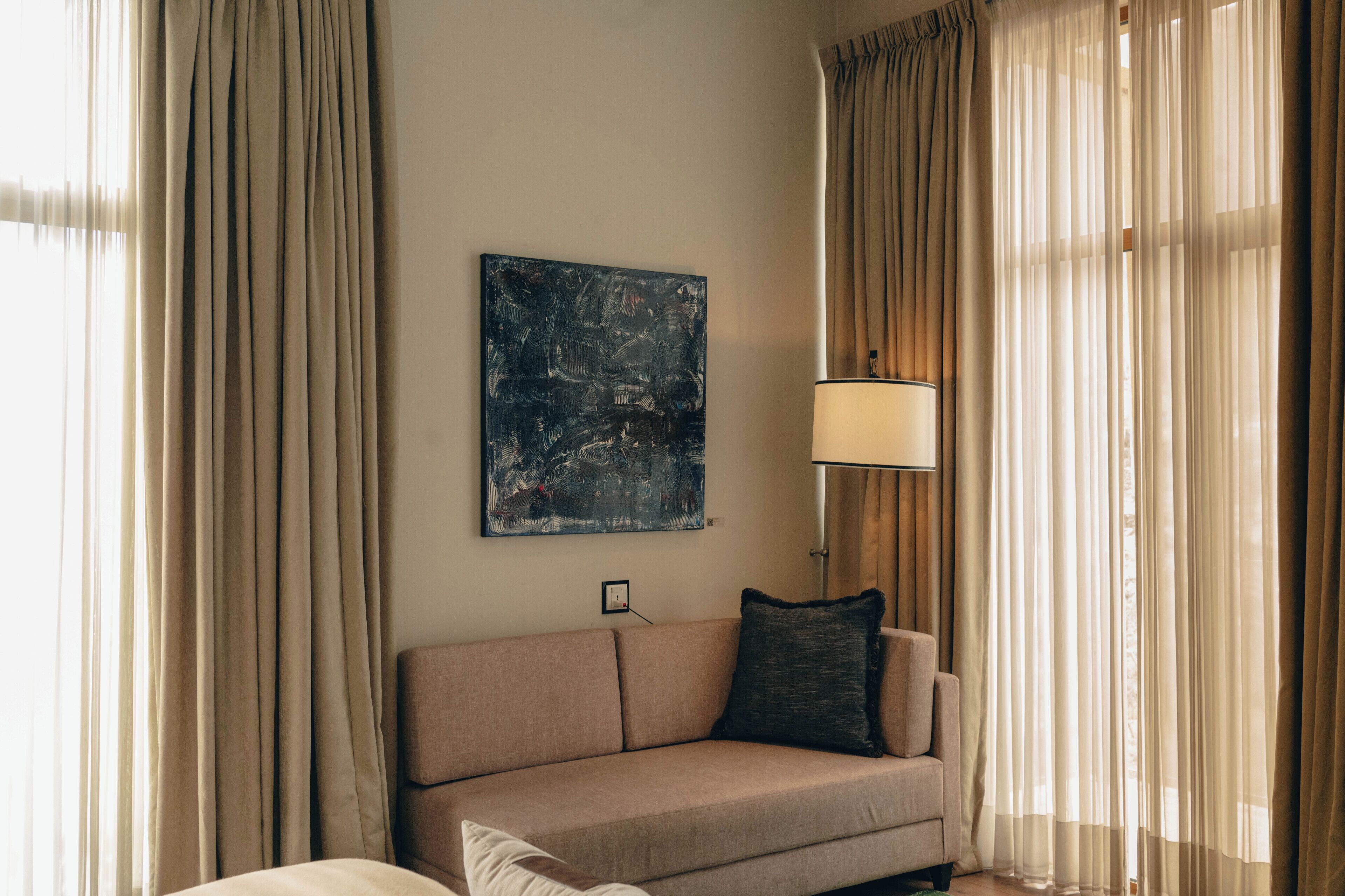 Uma sala de estar em tons neutros, com um sofá bege, uma pintura abstrata escura e cortinas translúcidas.