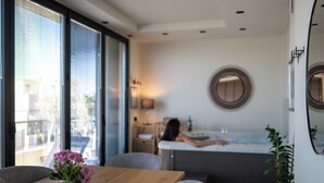 Quarto moderno com uma grande janela, uma mesa de madeira com um vaso de flores, um espelho redondo na parede e uma banheira com alguém relaxando.