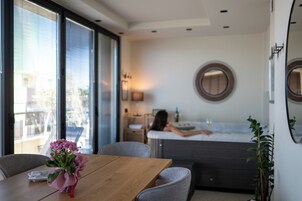 Quarto moderno com uma grande janela, uma mesa de madeira com um vaso de flores, um espelho redondo na parede e uma banheira com alguém relaxando.