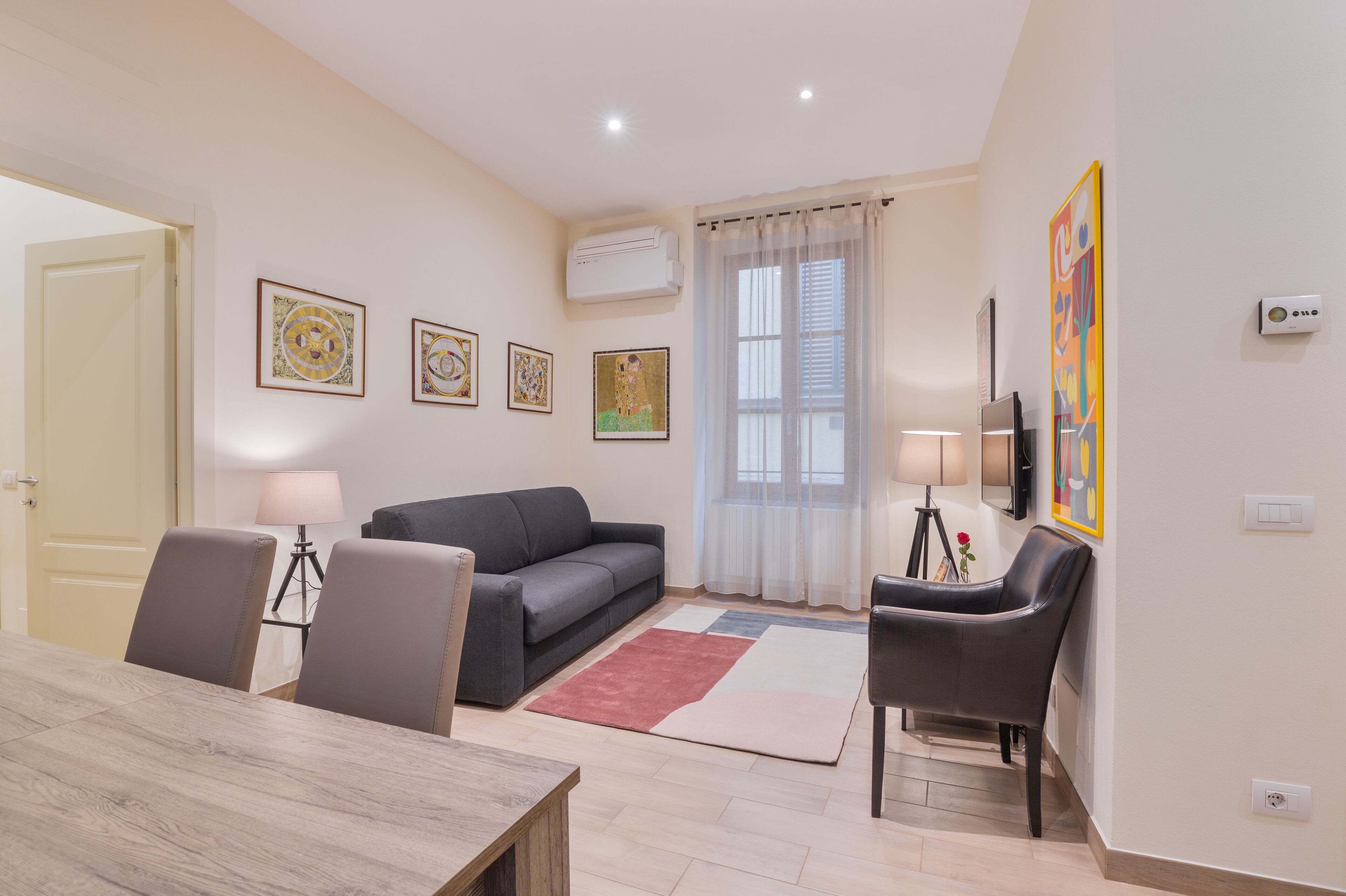 Uma sala de estar moderna com um sofá cinzento, uma mesa de jantar de madeira e uma TV fixada na parede.