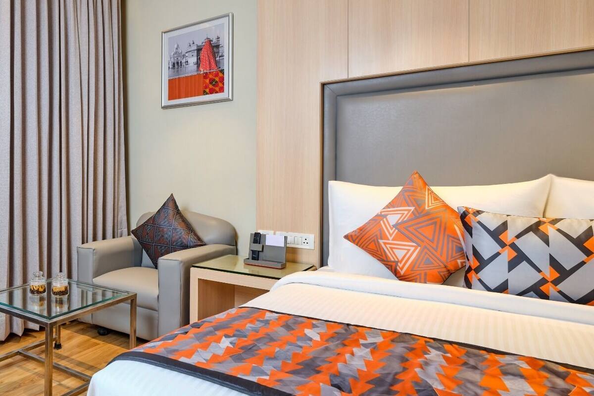 Quarto de hotel moderno com uma cama grande, uma mesa de vidro com uma cafeteira, uma cadeira e uma escrivaninha com um computador.