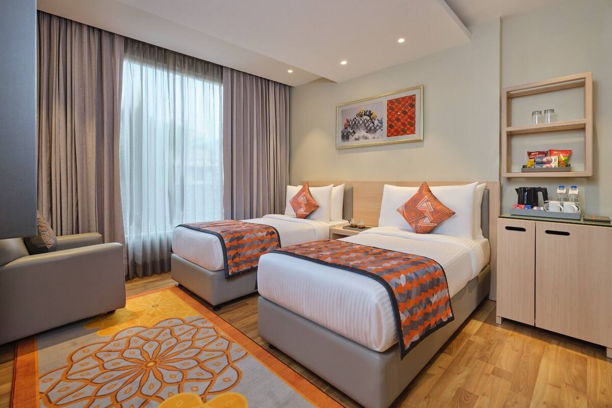 Quarto de hotel com duas camas, um sofá, uma mesa de centro, um minibar e uma janela grande com cortinas.