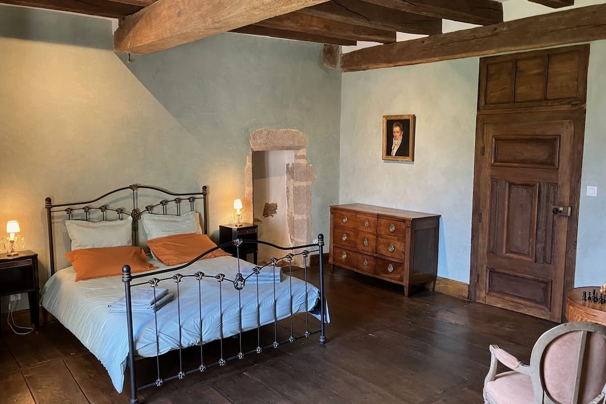 Quarto com cama de estrutura metálica, cômoda de madeira e uma grande porta de madeira.