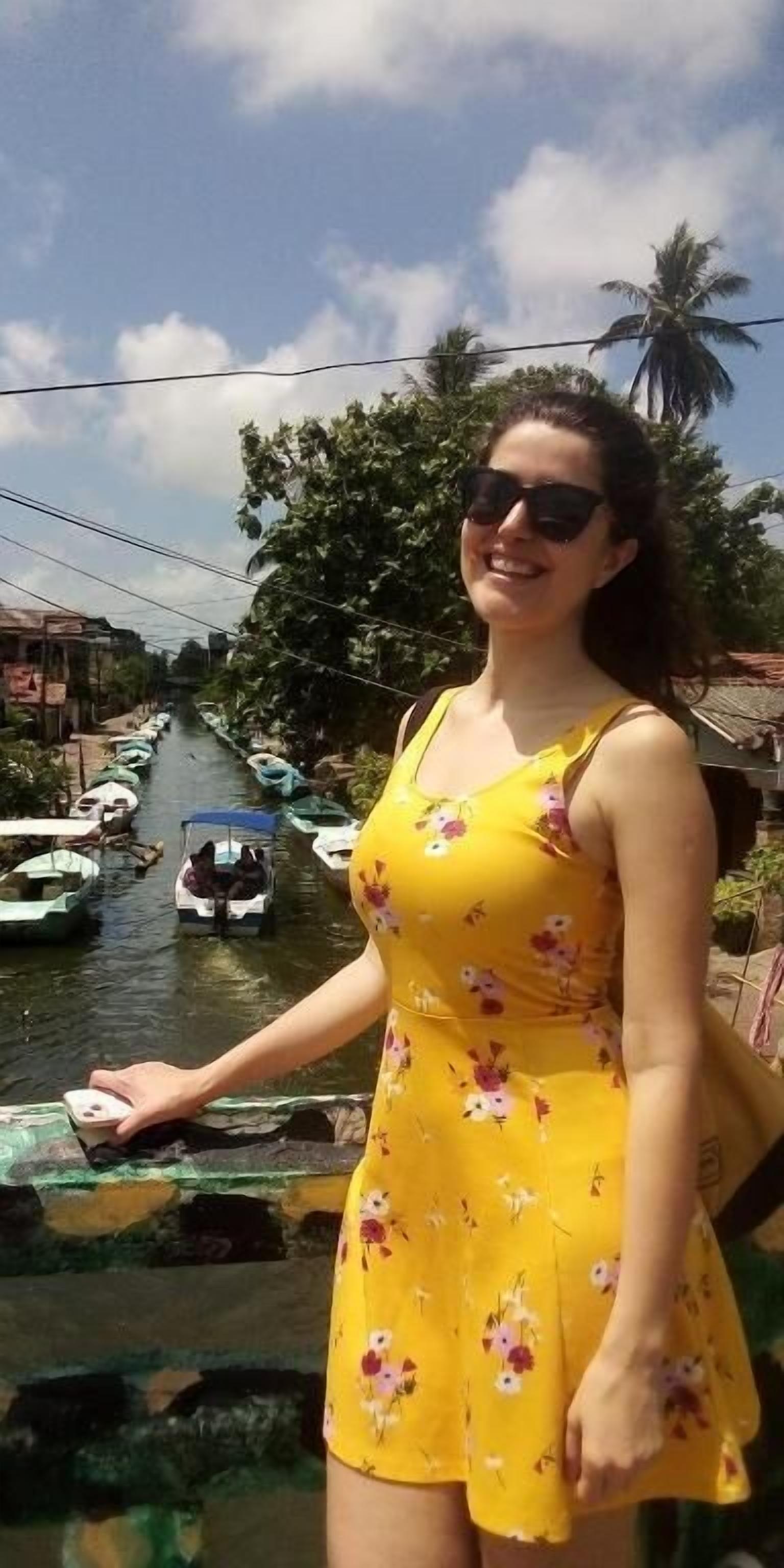 Mulher com vestido floral amarelo em uma ponte, com barcos em um canal.