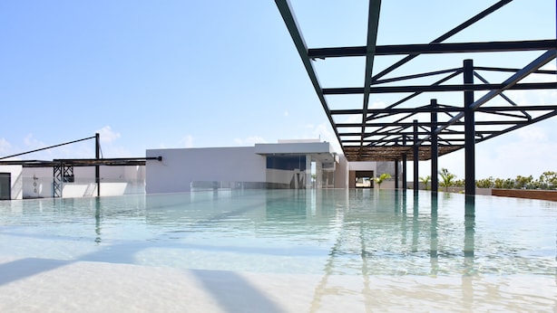 Uma área de piscina moderna com águas cristalinas, estrutura de cobertura em metal e um edifício branco ao fundo.