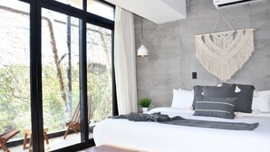 Quarto moderno com uma cama grande, um banquinho de madeira, um painel de parede em macramé e vista para o exterior.
