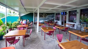 Um café ao ar livre com mesas e cadeiras, um mural de uma cascata e vista para uma ponte.
