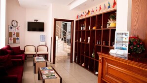 Um saguão de hotel com um sofá vermelho, duas poltronas, uma mesa de centro com revistas, uma estante de livros, uma televisão e um pinheiro de Natal.