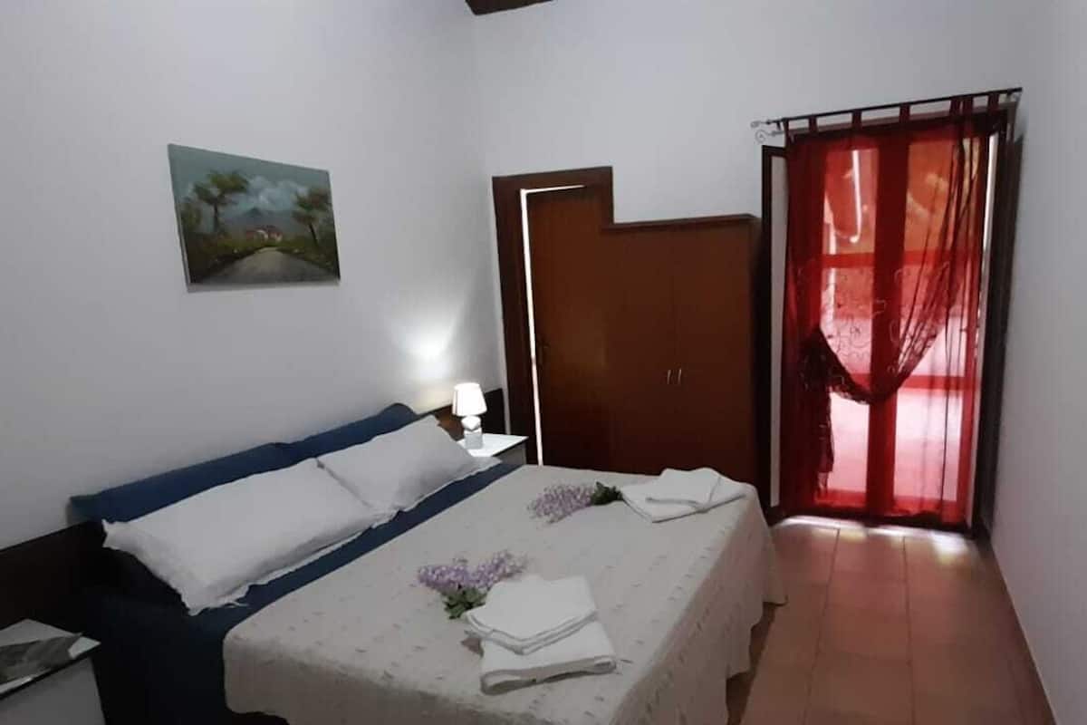 Quarto pequeno e limpo com uma cama, mesa de cabeceira, abajur, porta e janela com cortinas vermelhas.