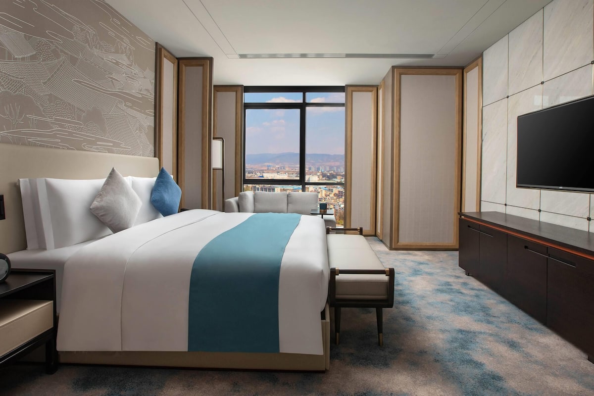 Quarto de hotel com uma cama grande, televisão, vista para a cidade e uma escrivaninha com cadeira.