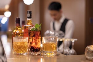 Três copos de bebidas cor de âmbar em um balcão de bar, com garrafas de licor ao fundo.